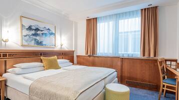 Quarto de hotel com cama grande, escrivaninha, cadeira, janela com cortinas, luminária de parede e um quadro.