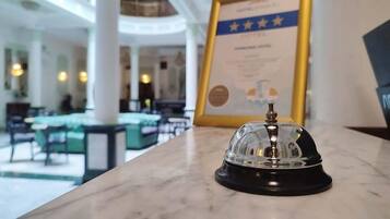 Uma área de recepção de hotel com um certificado de premiação emoldurado na parede, um sino sobre um balcão de mármore e uma visão parcial do saguão com mesas e cadeiras.
