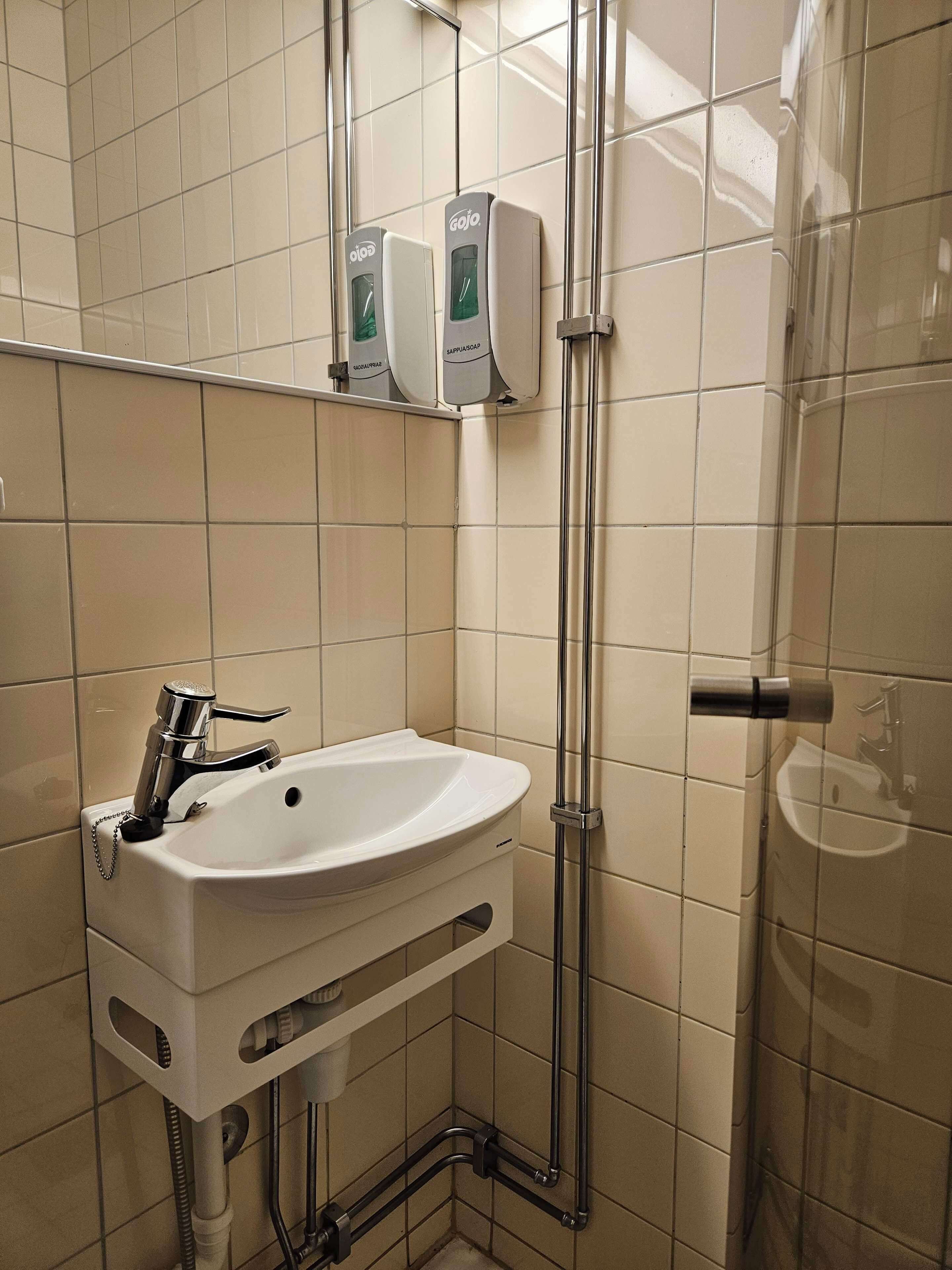 Casa de banho com lavatório branco, dispensador de sabonete manual e cabine de duche em vidro.