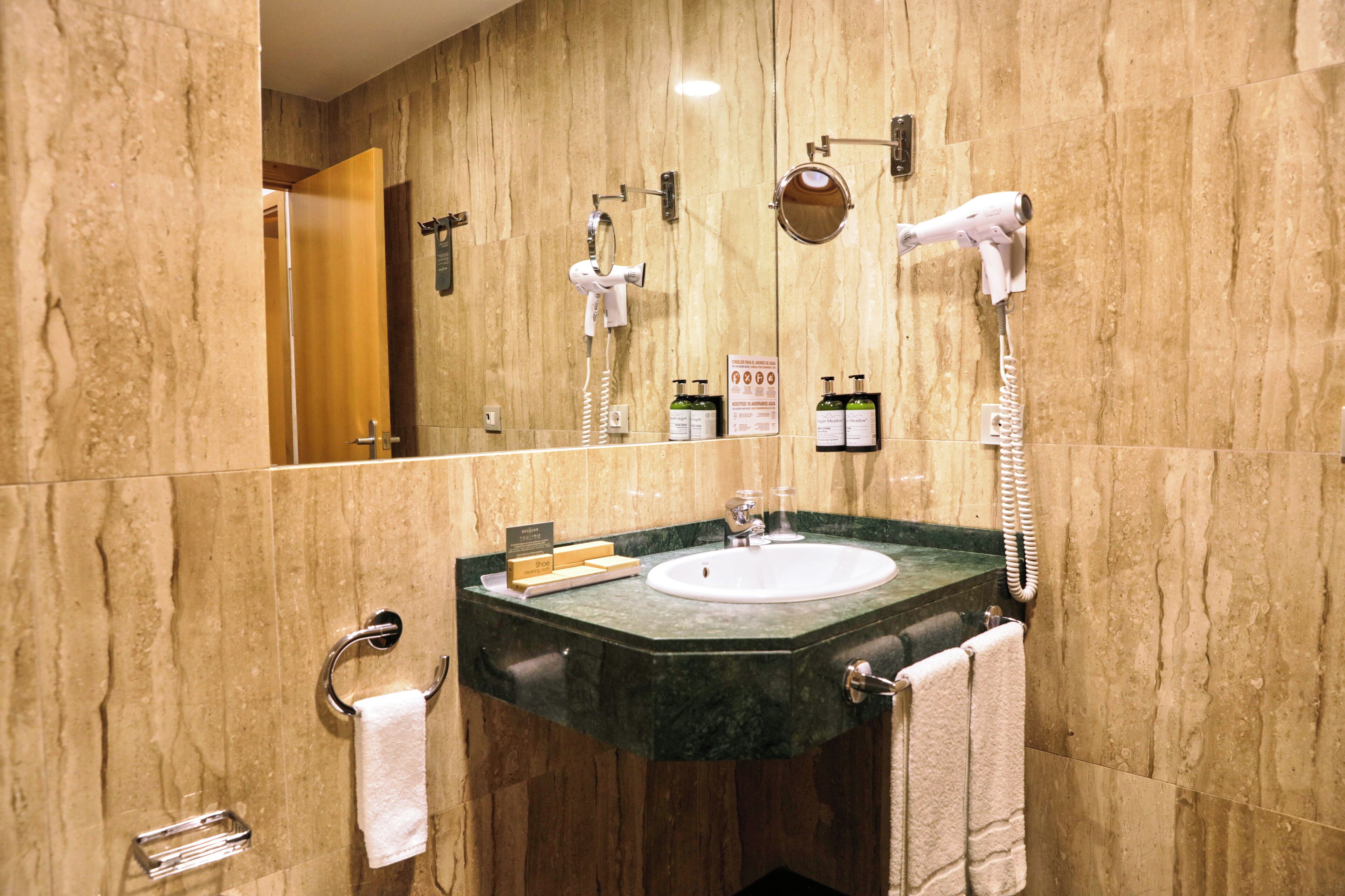 Uma casa de banho com parede de mármore, lavatório com espelho, porta-toalhas e secador de cabelo fixado na parede.