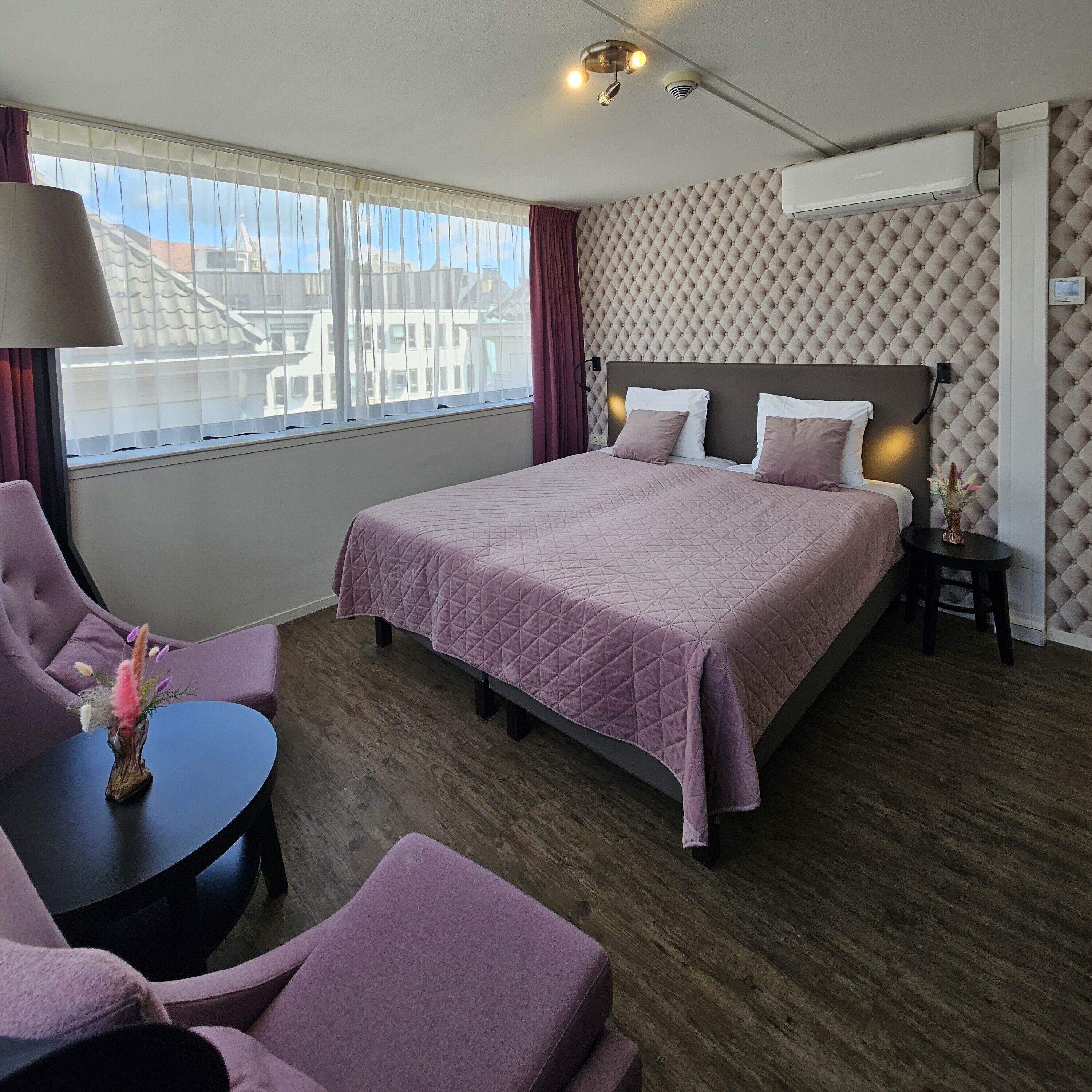 Quarto de hotel com uma cama grande, uma mesinha com um vaso e uma cadeira.