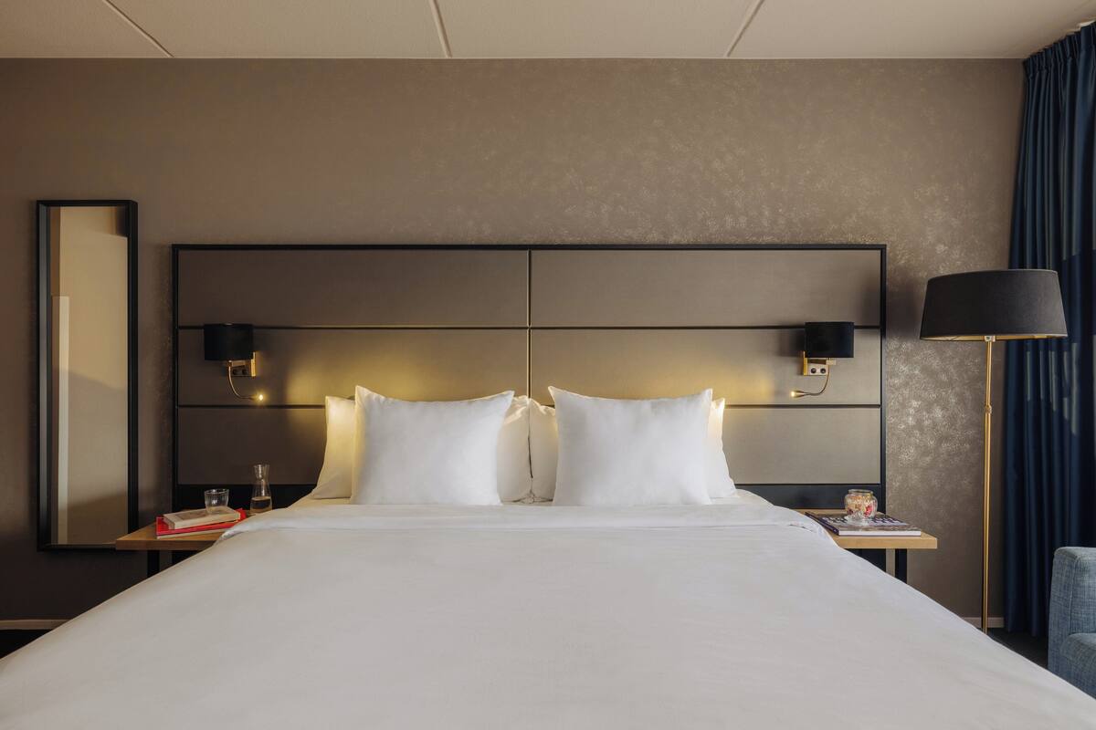 Quarto de hotel com uma cama grande, duas mesas de cabeceira, um criado-mudo, uma luminária e um espelho.