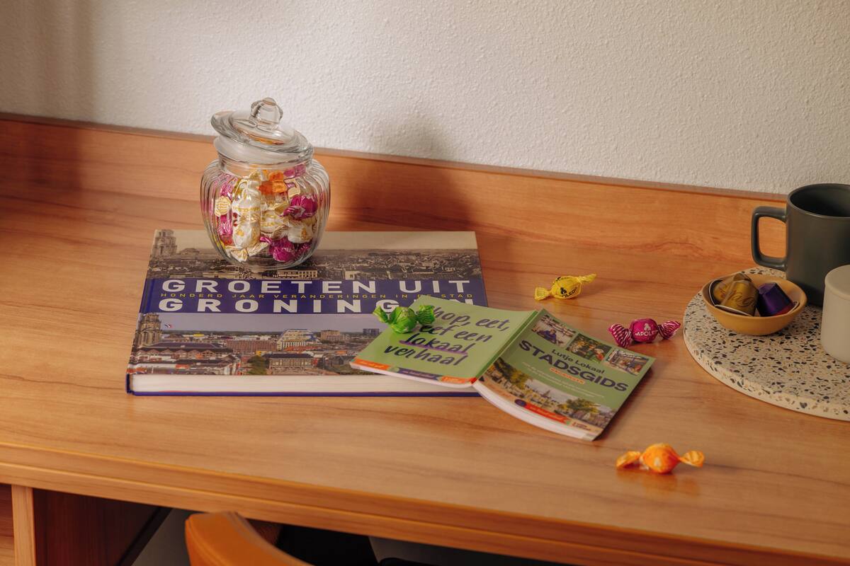 Uma mesa de madeira com um livro intitulado 'Groeten UIT GRONNING', um pote de vidro com doces, uma caneca de café e uma tigela de chocolates.