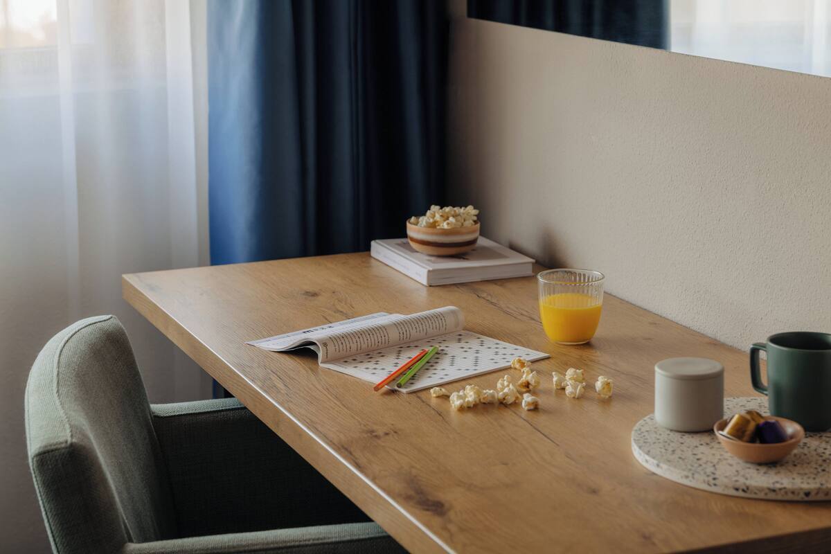 Uma mesa de madeira com um livro, um recipiente de pipocas, um copo de sumo de laranja, uma chávena verde e uma pequena tigela de petiscos.