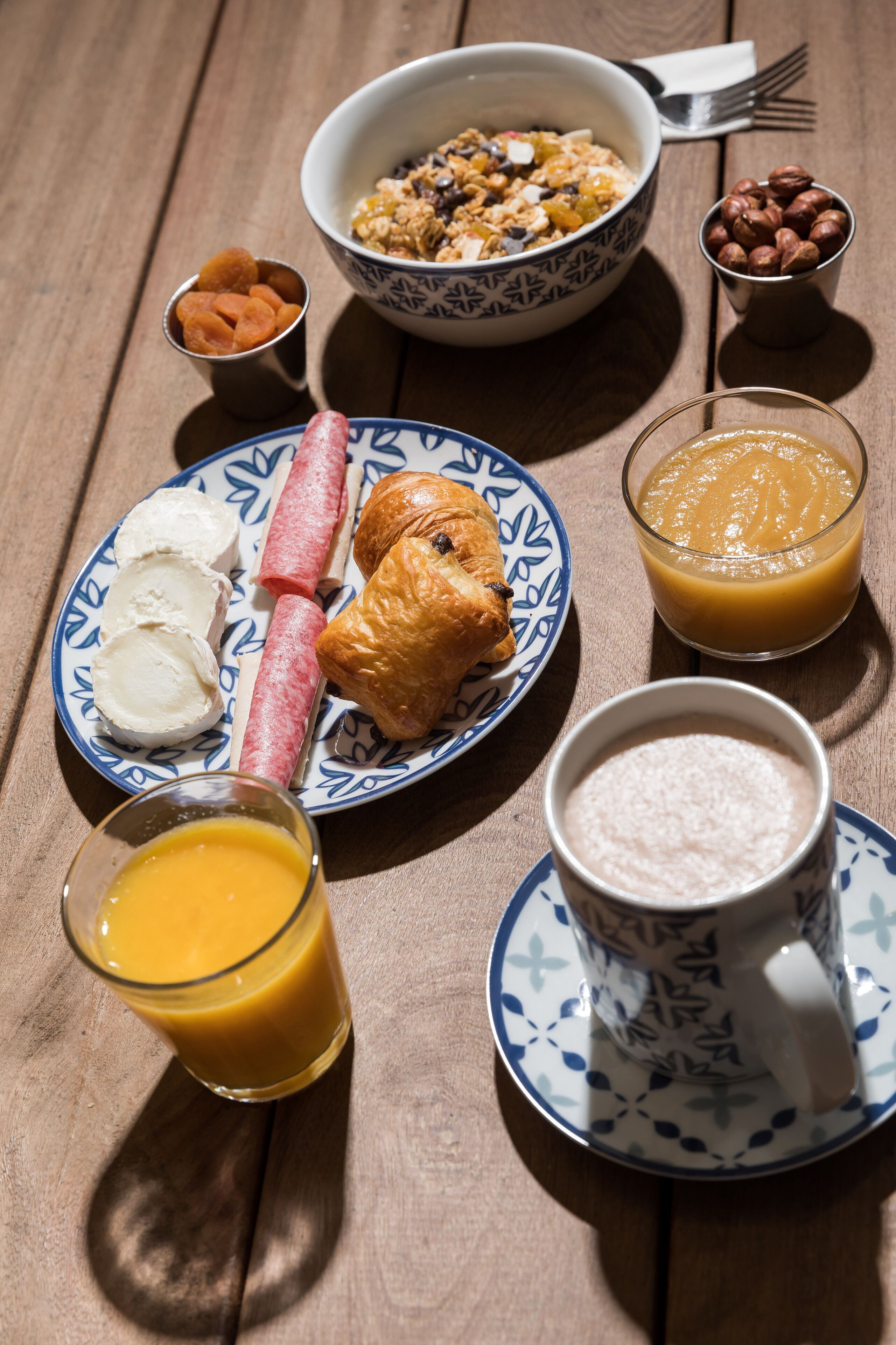 Uma mesa de madeira com uma bandeja de croissants, um copo de suco de laranja, uma tigela de cereais e uma xícara de café.
