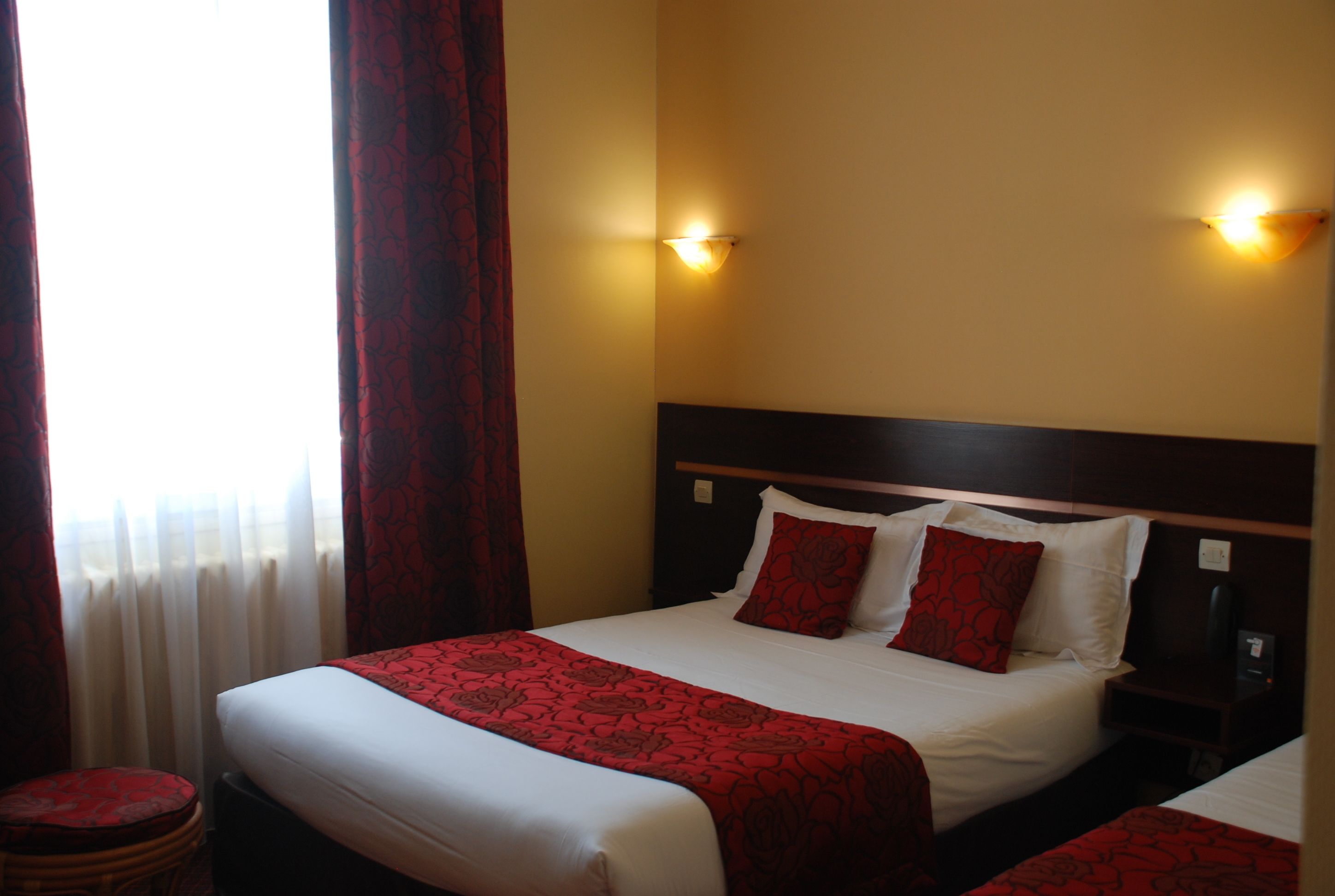 Quarto de hotel com uma cama grande, duas mesas de cabeceira, um cobreleite com padrão vermelho e uma janela com cortinas.