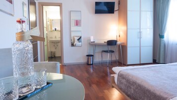 Quarto de hotel com uma cama, escrivaninha, cadeira, televisão presa na parede e banheiro visível por uma porta aberta.