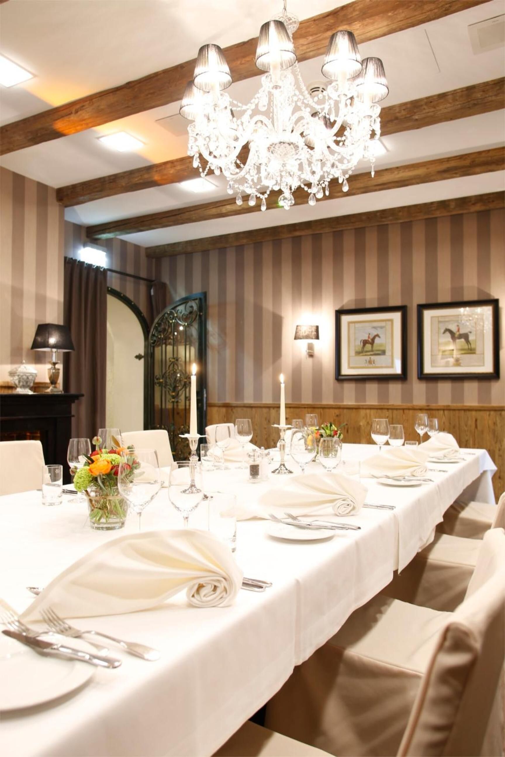 Uma sala de jantar com uma longa mesa posta para refeição, com um candelabro, quadros emoldurados e uma vela sobre a mesa.