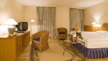 Quarto de hotel com cama, duas cadeiras, escrivaninha com televisão e mesinha com um vaso de flores.