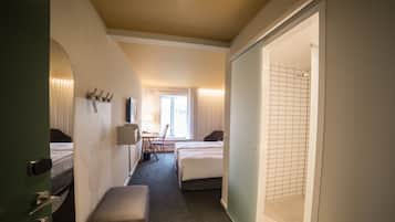 Quarto de hotel com duas camas, uma escrivaninha, uma cadeira, uma luminária e banheiro com chuveiro.