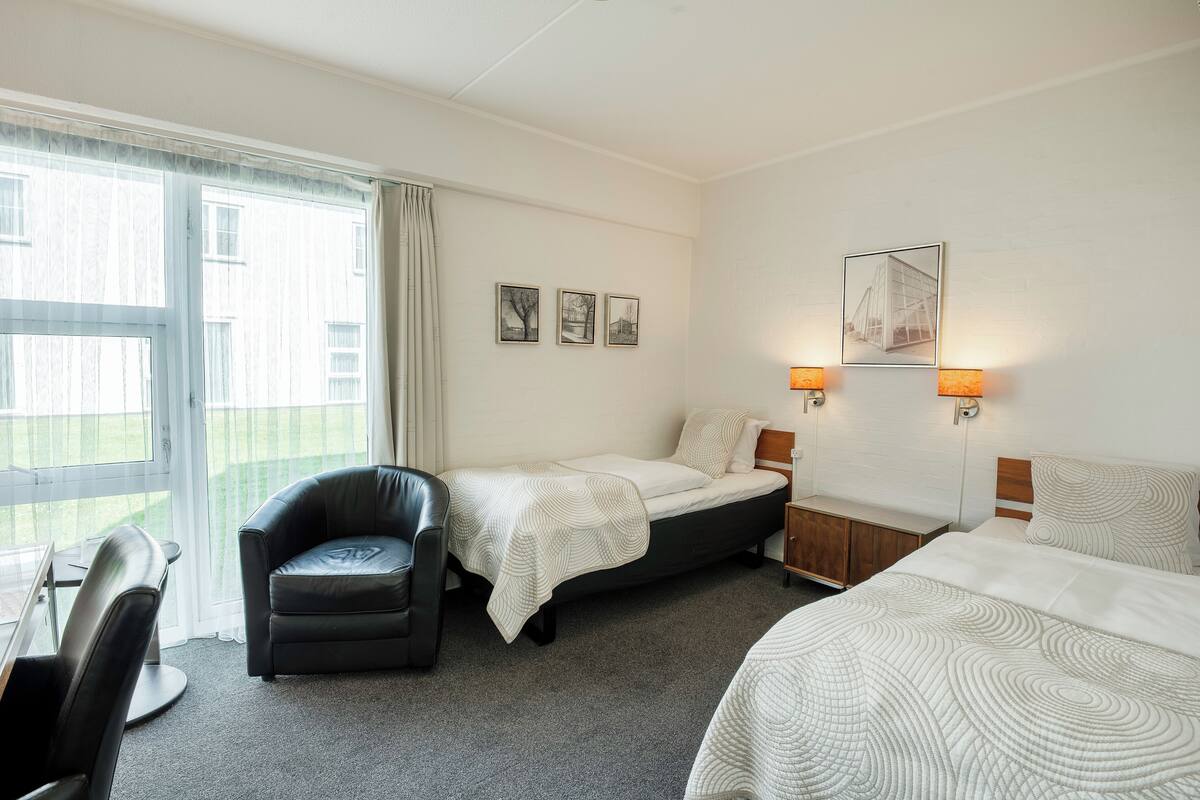 Quarto de hotel com cama, cadeira, escrivaninha e janela com cortinas. Há três quadros na parede e duas luminárias.