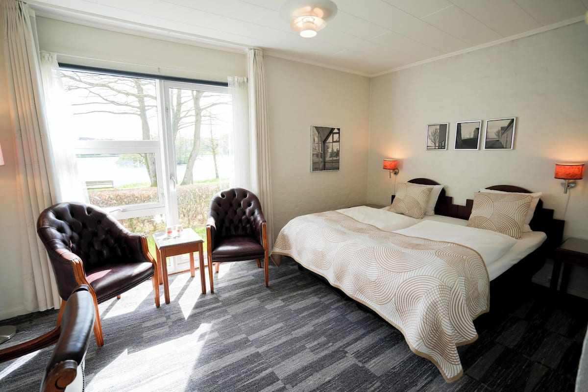Quarto de hotel com uma cama grande, duas poltronas, uma mesa pequena e uma janela com cortinas.