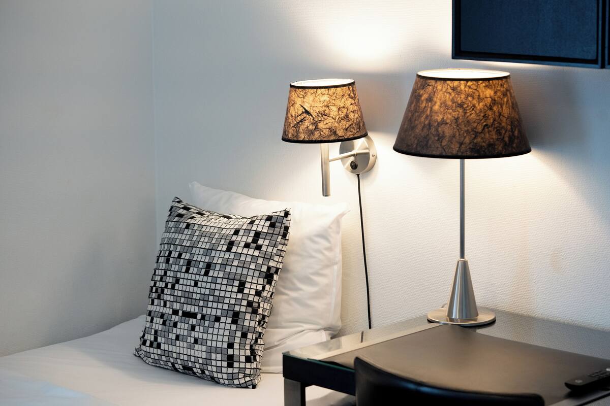 Cama bem arrumada com almofada estampada, mesa de cabeceira com abajur e televisão fixada na parede.