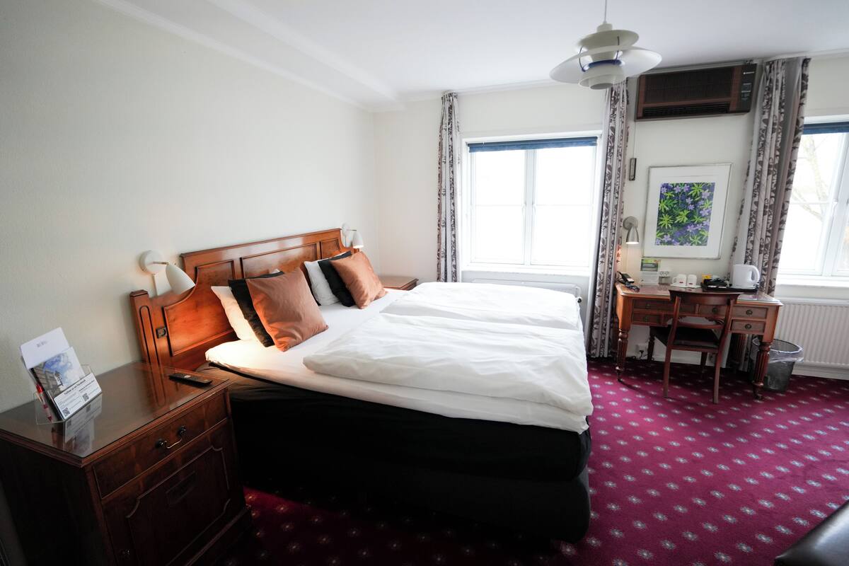 Quarto de hotel com cama, escrivaninha, cadeira, abajur e janela.