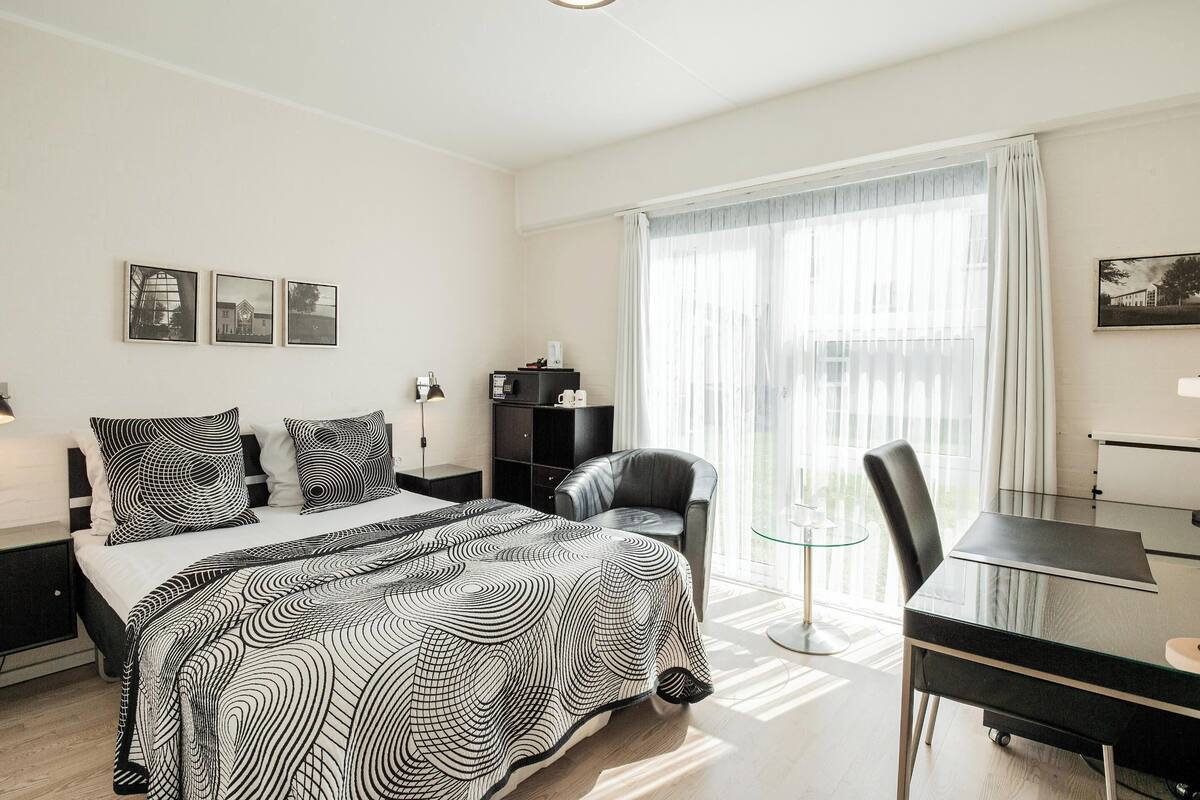 Quarto moderno com uma cama grande, uma escrivaninha e uma cadeira. Há três quadros na parede, um guarda-roupa e uma janela com cortinas.