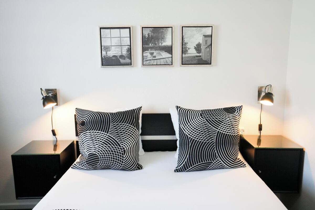 Uma cama bem arrumada com almofadas estampadas em preto e branco, ladeada por duas mesas de cabeceira pretas com abajures, e três quadros emoldurados a preto e branco na parede acima.