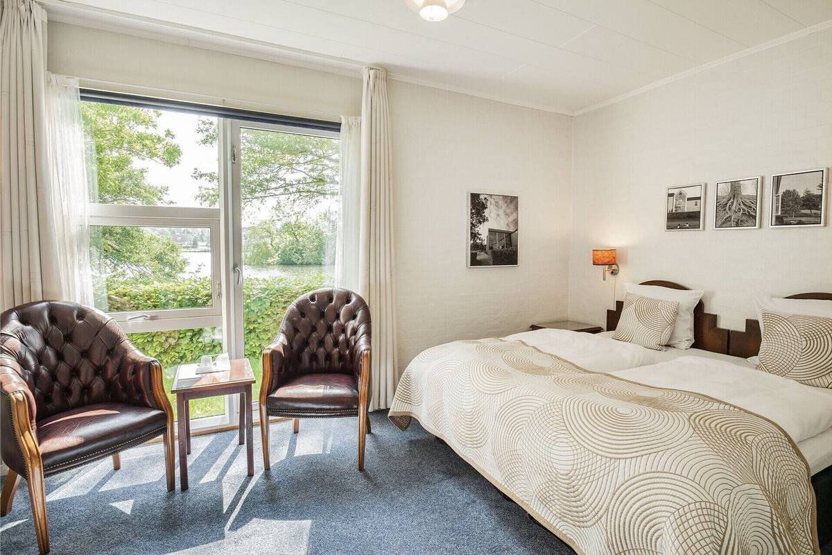 Quarto de hotel com uma cama grande, duas poltronas, uma mesa pequena e uma janela com vista para o verde.