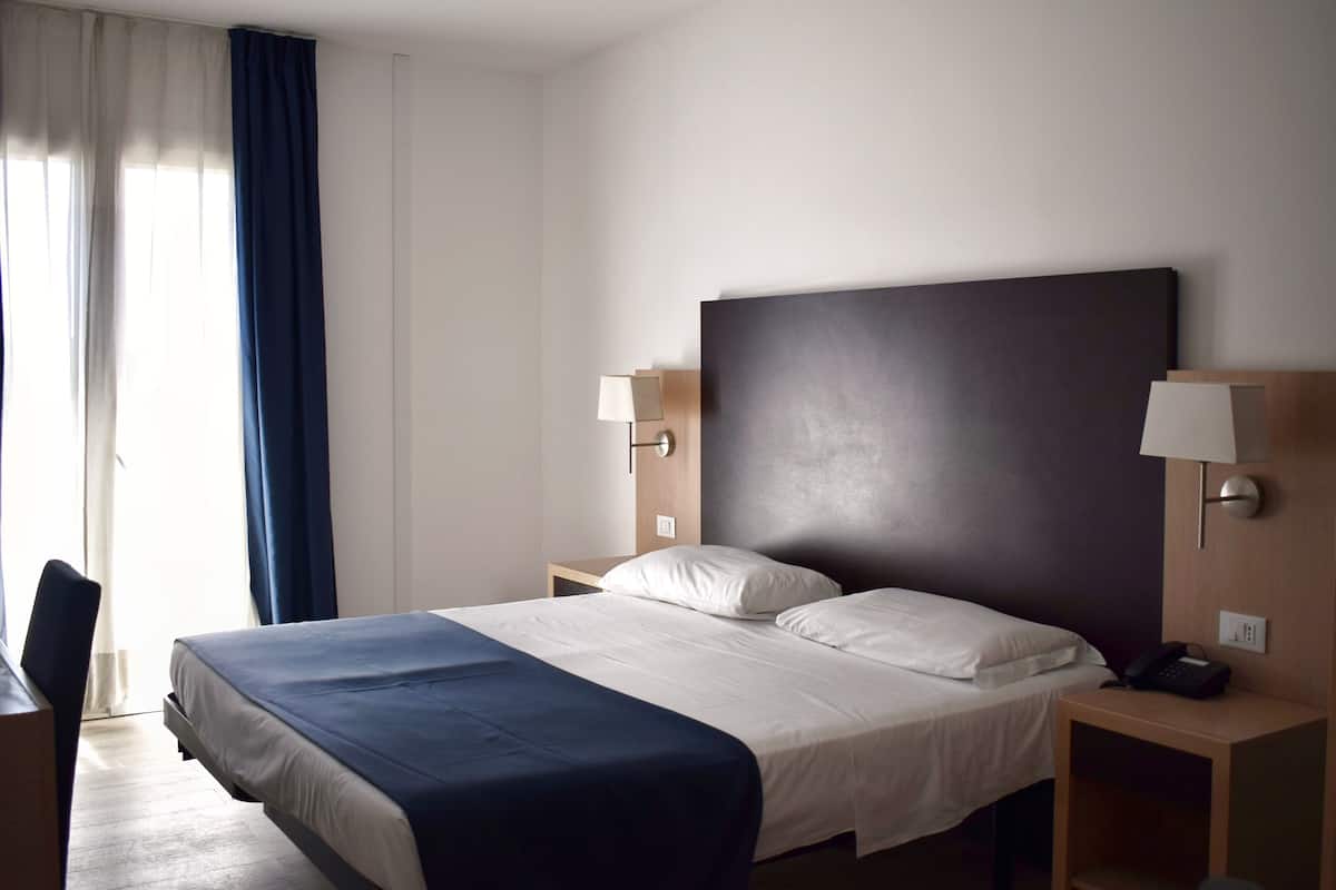 Quarto de hotel com uma cama grande, duas mesas de cabeceira, uma cadeira e uma janela com cortinas.