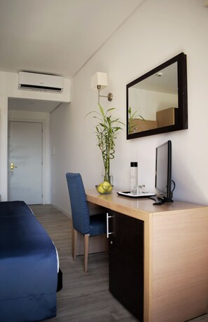 Quarto de hotel com cama, escrivaninha com computador, cadeira, espelho, planta em vaso e ar condicionado.