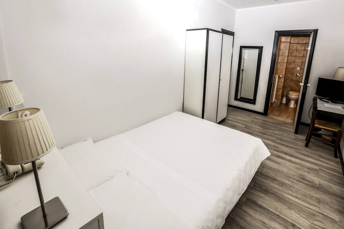 Quarto de hotel com cama, secretária com luminária, guarda-roupa e banheiro visível por uma porta aberta.