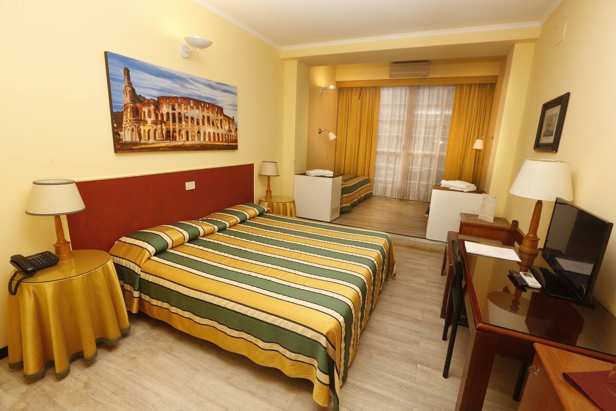 Quarto de hotel com cama, escrivaninha, televisão e um quadro de um edifício histórico.