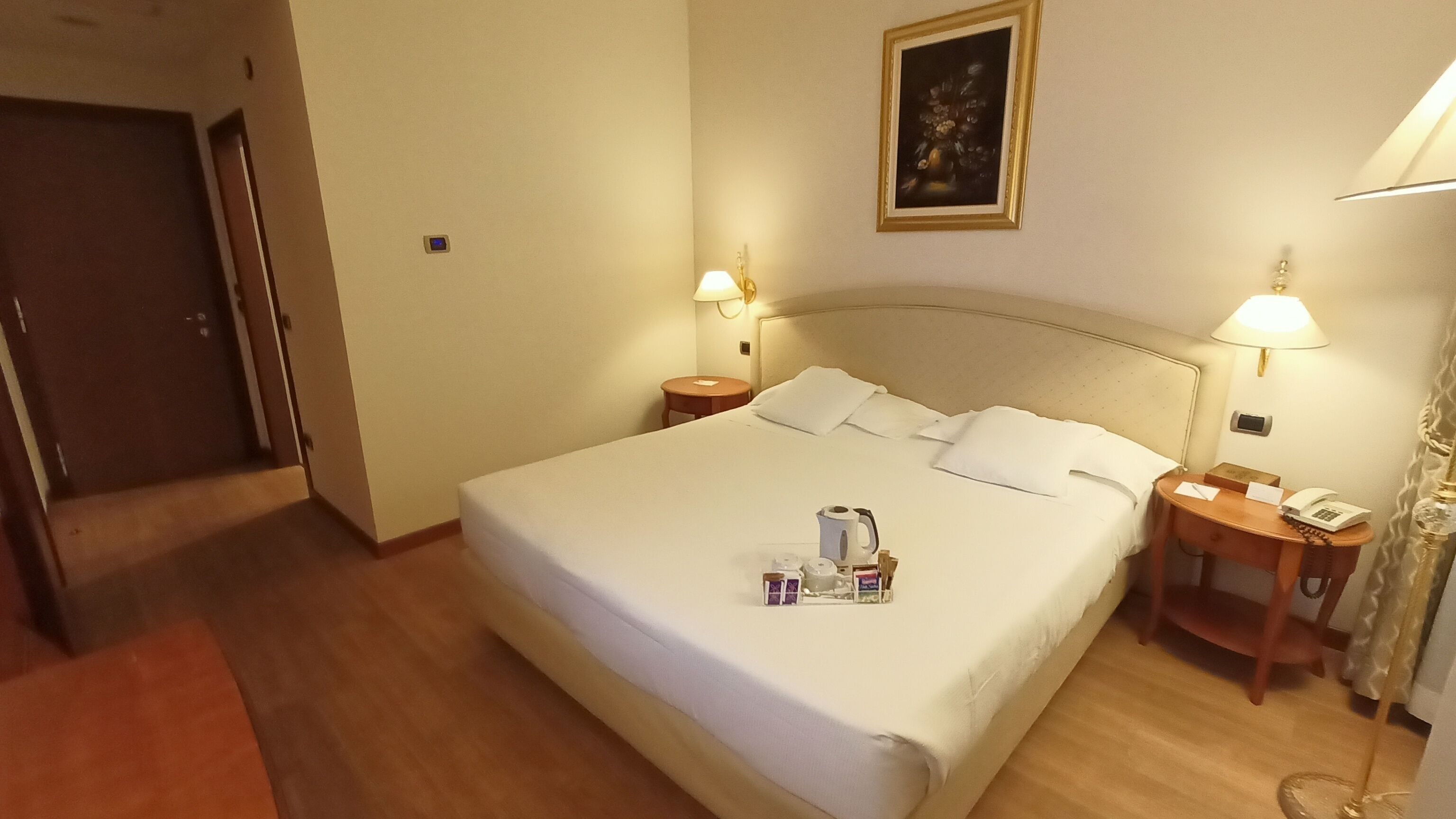 Quarto de hotel com uma cama grande, duas mesas de cabeceira com luminárias, um quadro na parede e uma porta que dá para outro quarto.