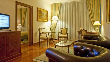 Quarto de hotel com armário de madeira, sofá de couro, mesa de centro de vidro, duas poltronas, uma mesinha de cabeceira com abajur e uma televisão exibindo uma página da web.