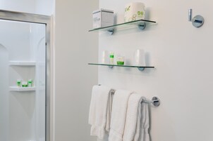 Casa de banho com cabine de chuveiro em vidro, toalheiro branco com toalhas e estantes com produtos de higiene pessoal.