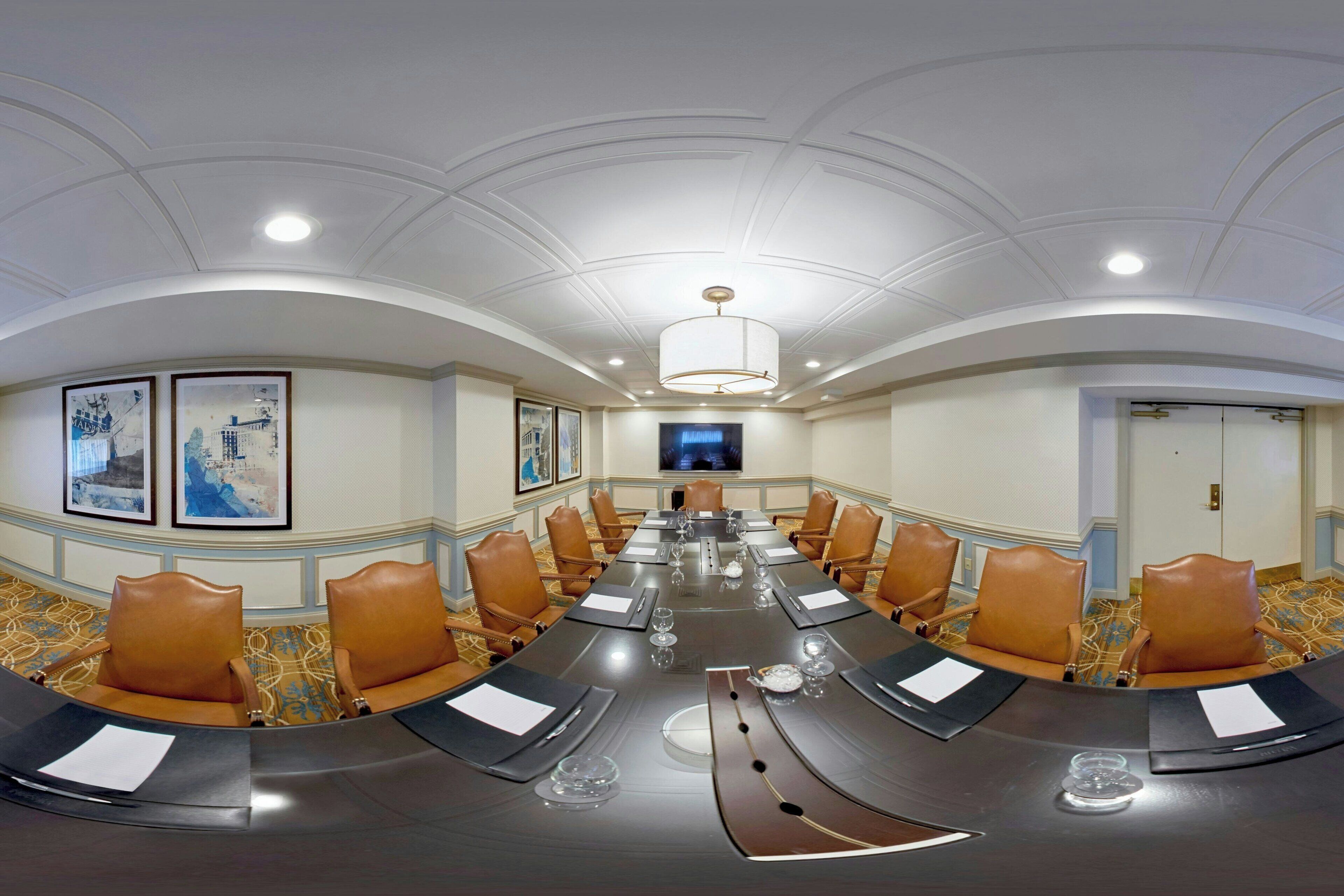 Uma sala de conferência com uma mesa comprida, cadeiras de couro marrom, uma televisão de tela plana e obras de arte emolduradas nas paredes.