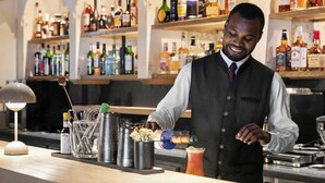 Um bartender preparando uma bebida num bar, com prateleiras cheias de garrafas de licor ao fundo.