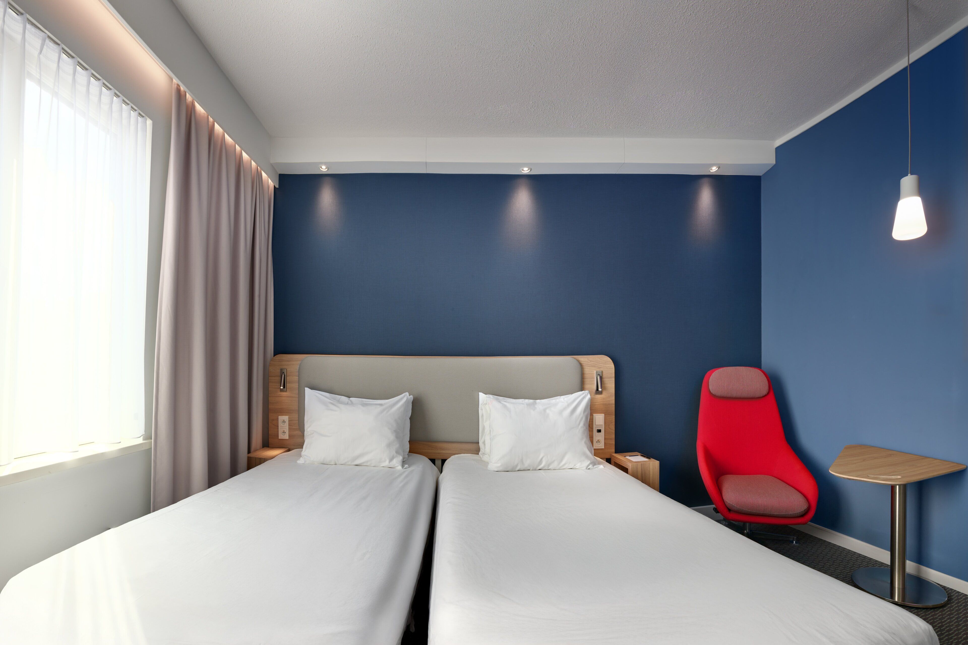 Quarto de hotel com duas camas, uma cadeira vermelha, uma mesinha e uma parede azul.