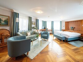Quarto de hotel moderno com sofá, poltrona e cama. Há uma mesa de trabalho com cadeira, luminária e um vaso com flores.