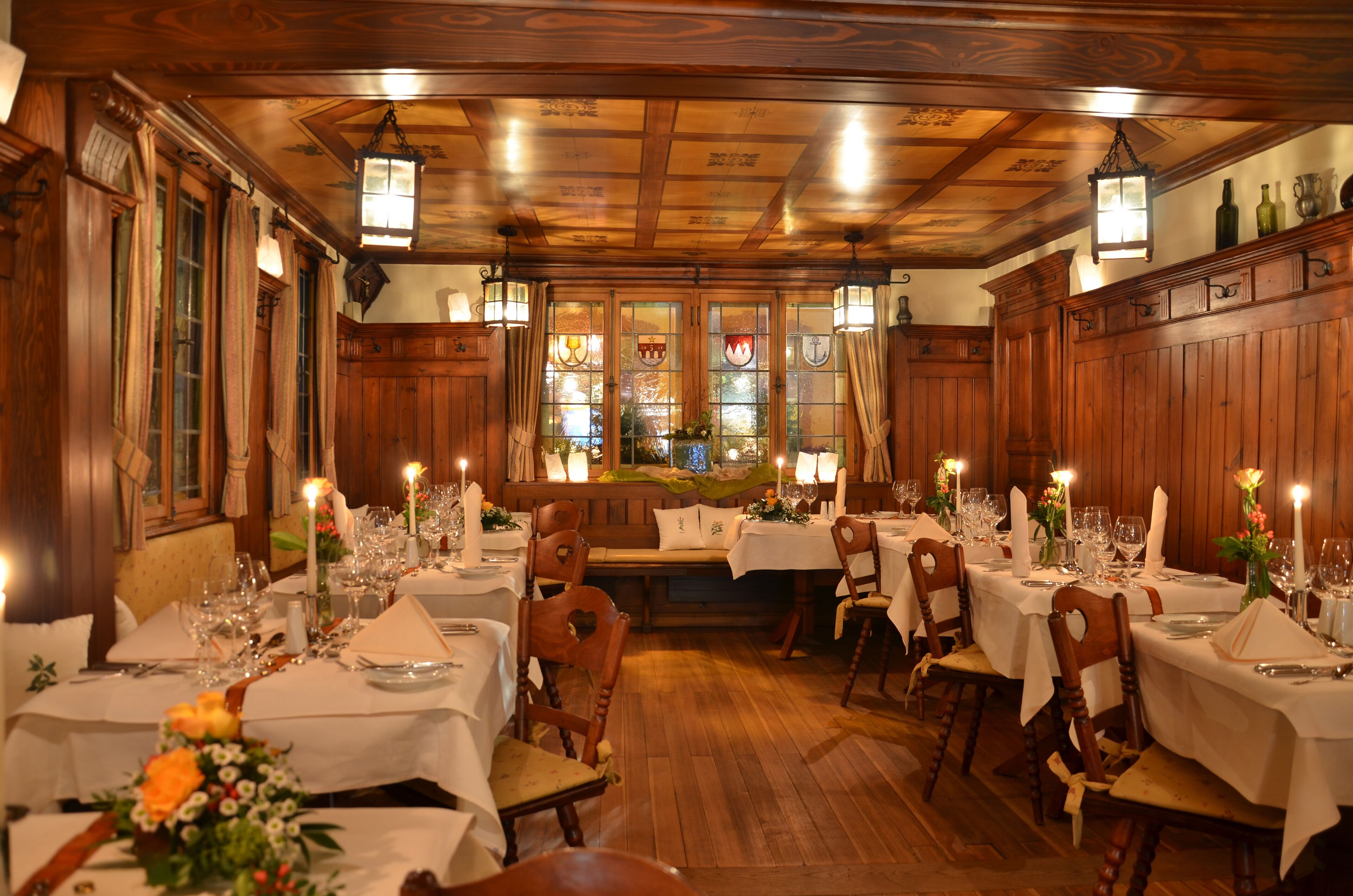 Uma sala de jantar com painéis de madeira, toalhas brancas, mesas iluminadas por velas e um banco.