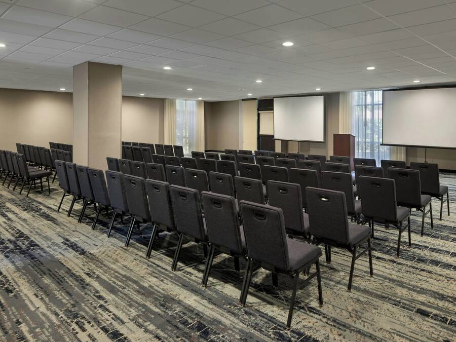 Uma sala de conferências com fileiras de cadeiras, piso coberto por carpete e uma tela de projeção.