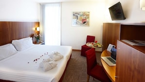Quarto de hotel com uma cama grande, uma secretária com um laptop, uma cadeira, uma mesa pequena com comida e uma TV presa à parede.