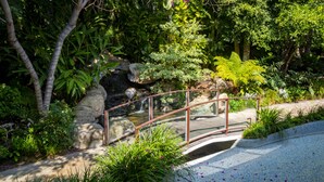 Um jardim com uma pequena ponte sobre um espelho d'água, cercado por vegetação exuberante e plantas floridas.