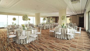 Um amplo salão de banquetes com mesas e cadeiras redondas, tetos altos e grandes janelas.