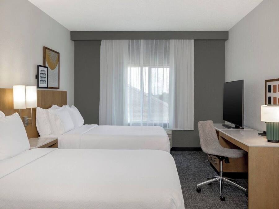 Quarto de hotel com duas camas, uma escrivaninha, uma cadeira, uma televisão e uma janela com cortinas.