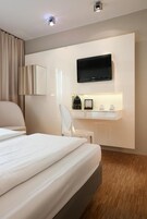 Quarto de hotel moderno com uma cama grande, uma escrivaninha com cadeira, uma TV de tela plana na parede e uma mesa de cabeceira com abajur e relógio.