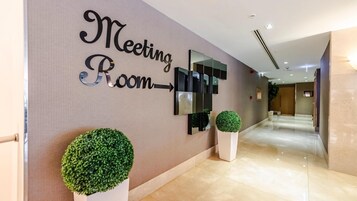 Corredor com placa indicando uma sala de reuniões, um espelho e dois vasos de plantas.