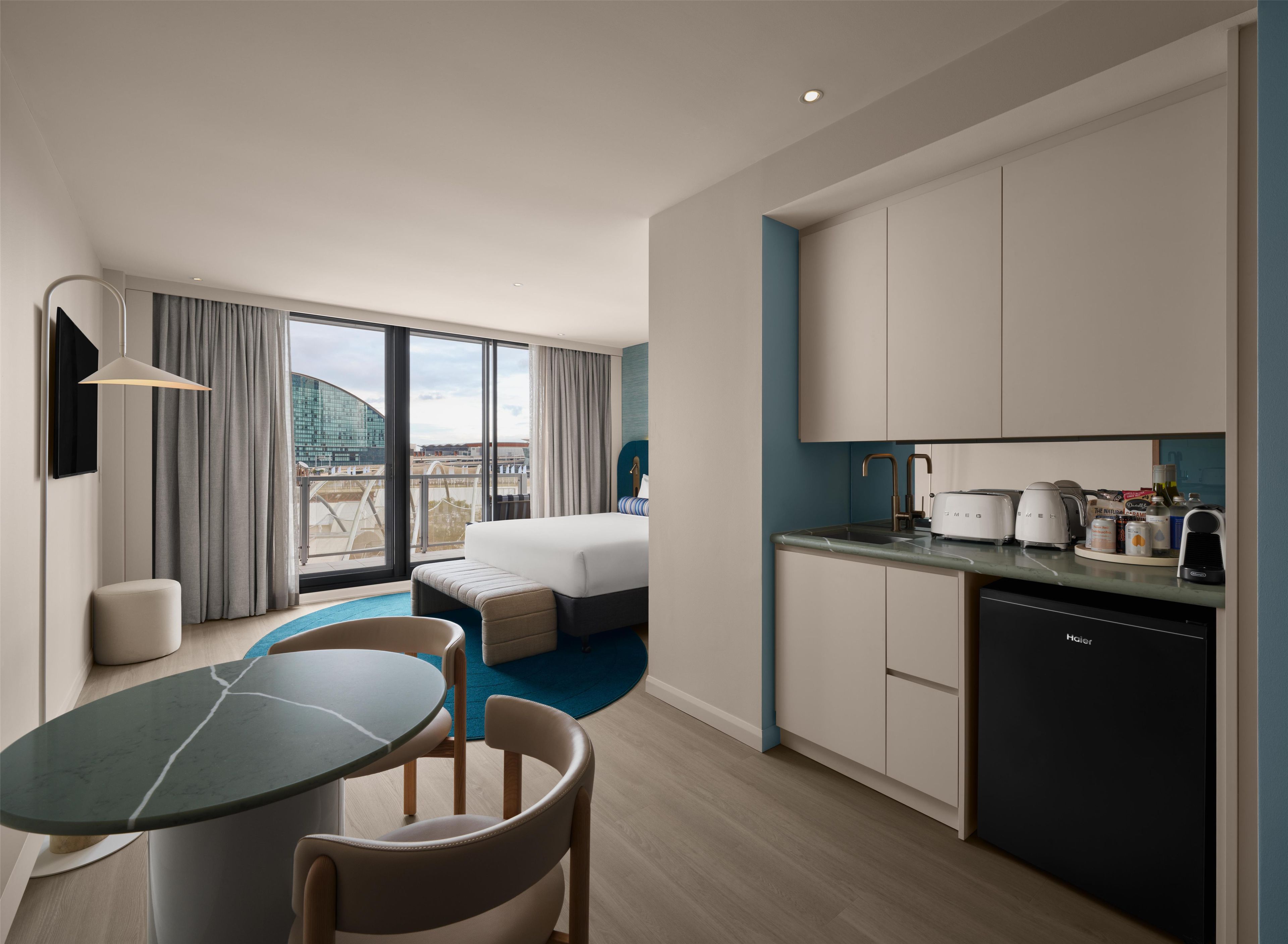 Quarto de hotel moderno com cama, mesa de jantar, cozinha e vista para a paisagem urbana.