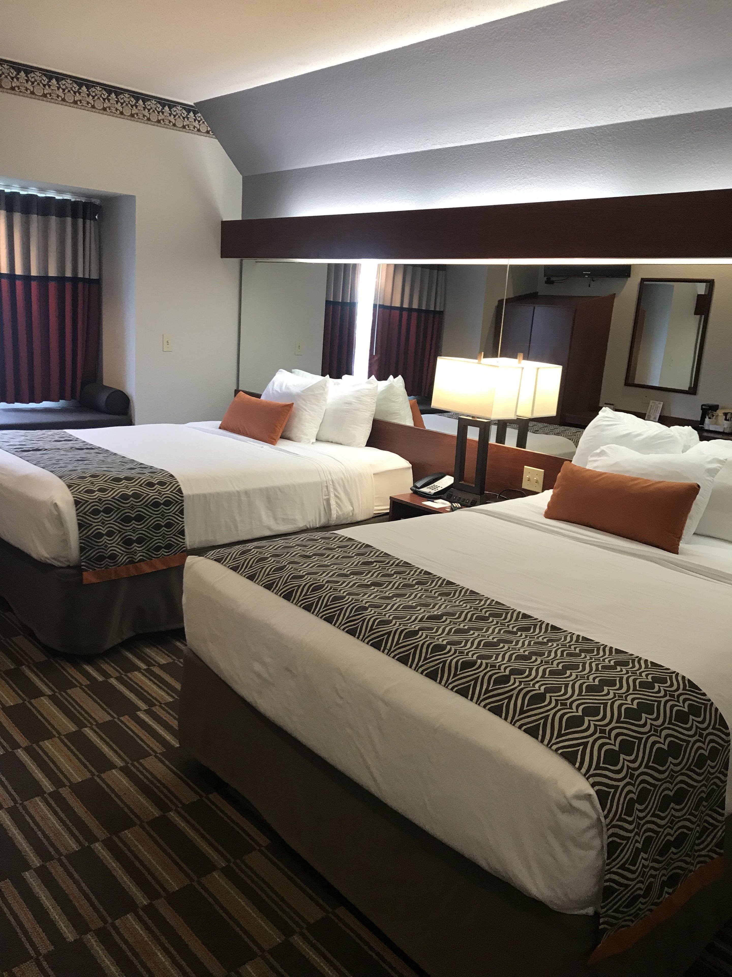 Quarto de hotel com duas camas, um espelho grande, uma luminária e uma janela com cortinas.