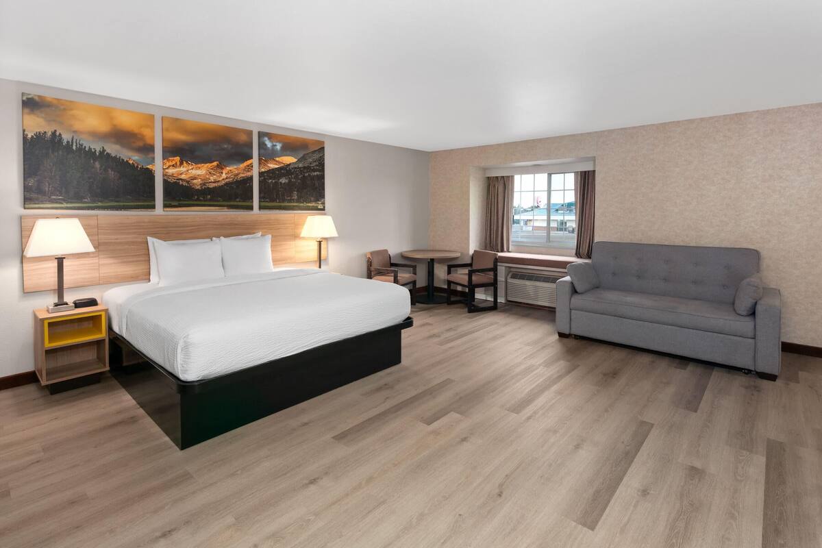 Quarto de hotel com cama, sofá cinza, mesa pequena, duas luminárias e janela com cortinas.