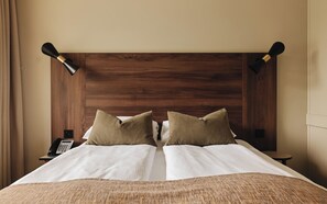 Uma cama de casal com duas almofadas, cabeceira de madeira, mesas de cabeceira com luminárias e um telefone em uma mesinha de apoio.