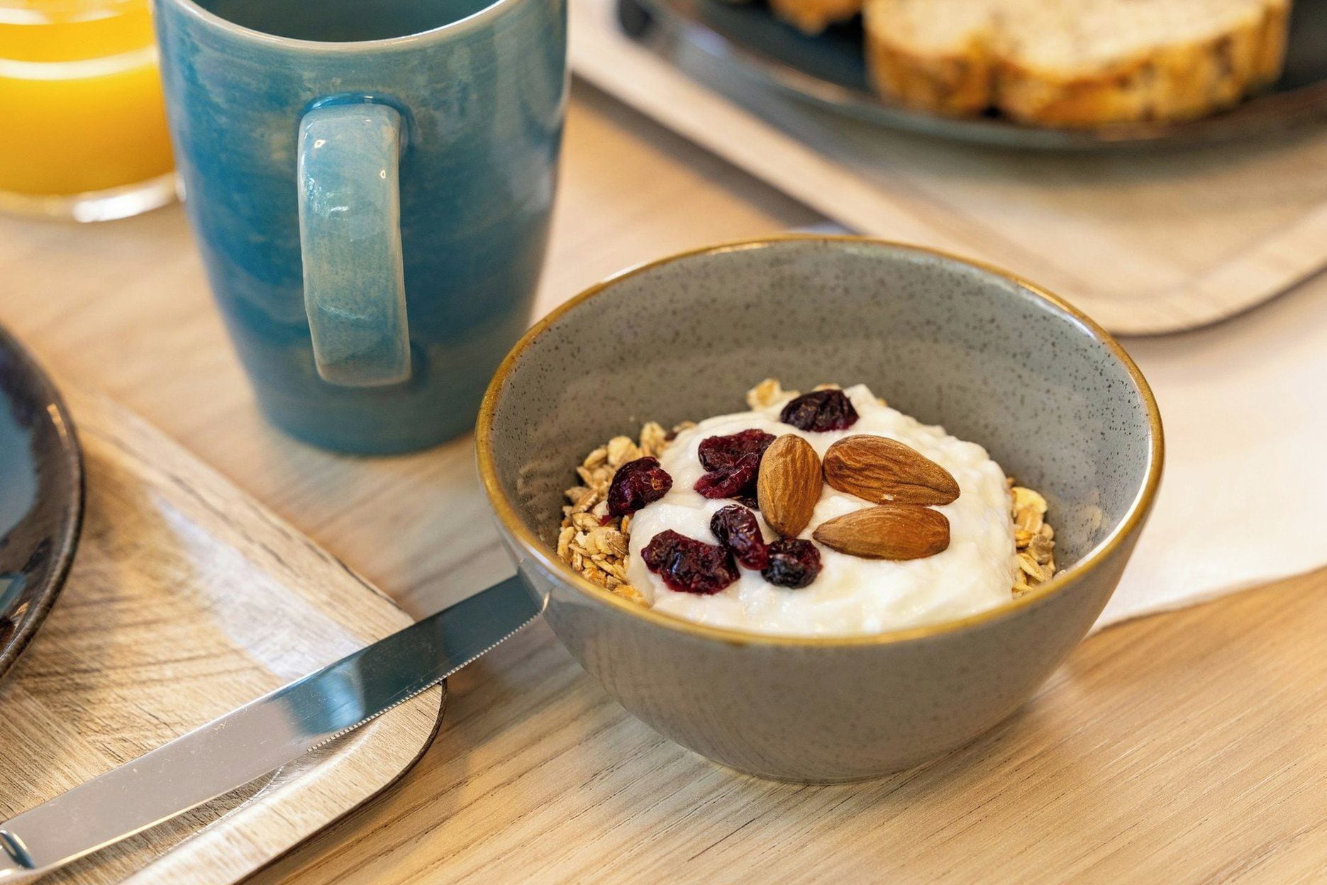 Mesa de café da manhã com tigela de cereal coberto de amêndoas e passas, caneca azul e pão torrado em um prato.