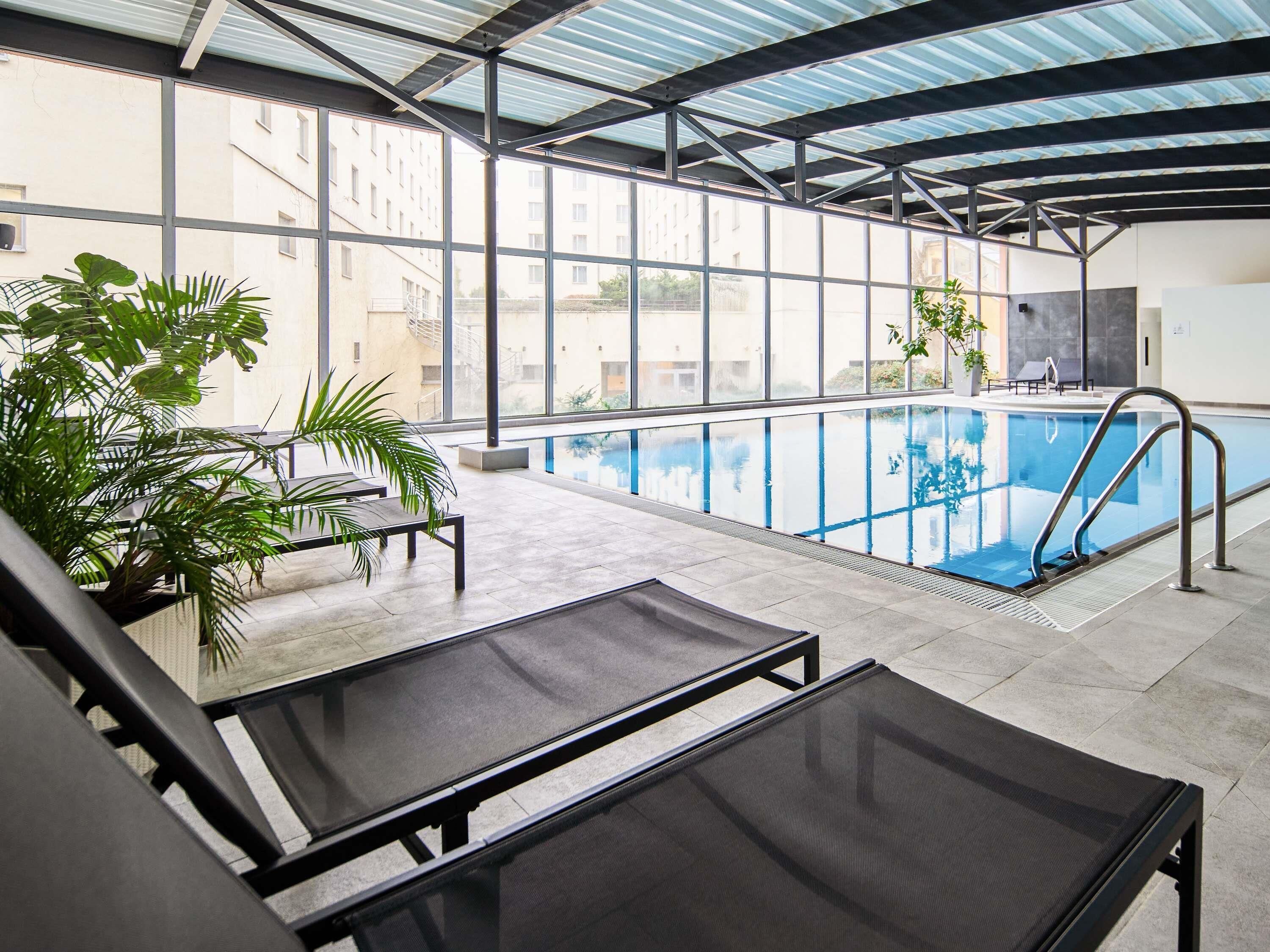 Piscina interior com paredes de vidro transparente, teto metálico e espreguiçadeiras.