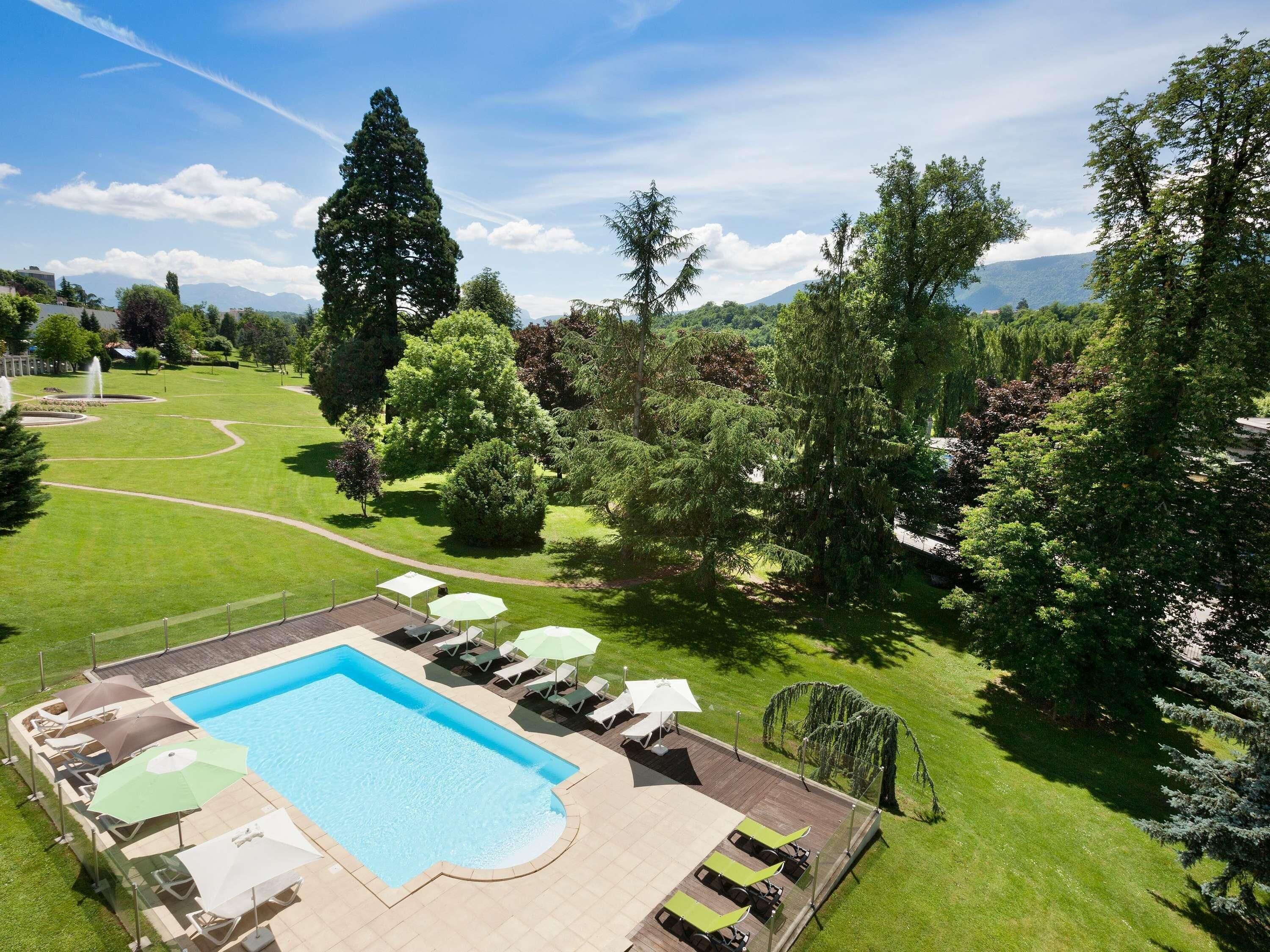 Piscina com espreguiçadeiras e guarda-sóis, cercada por um gramado bem cuidado e árvores.