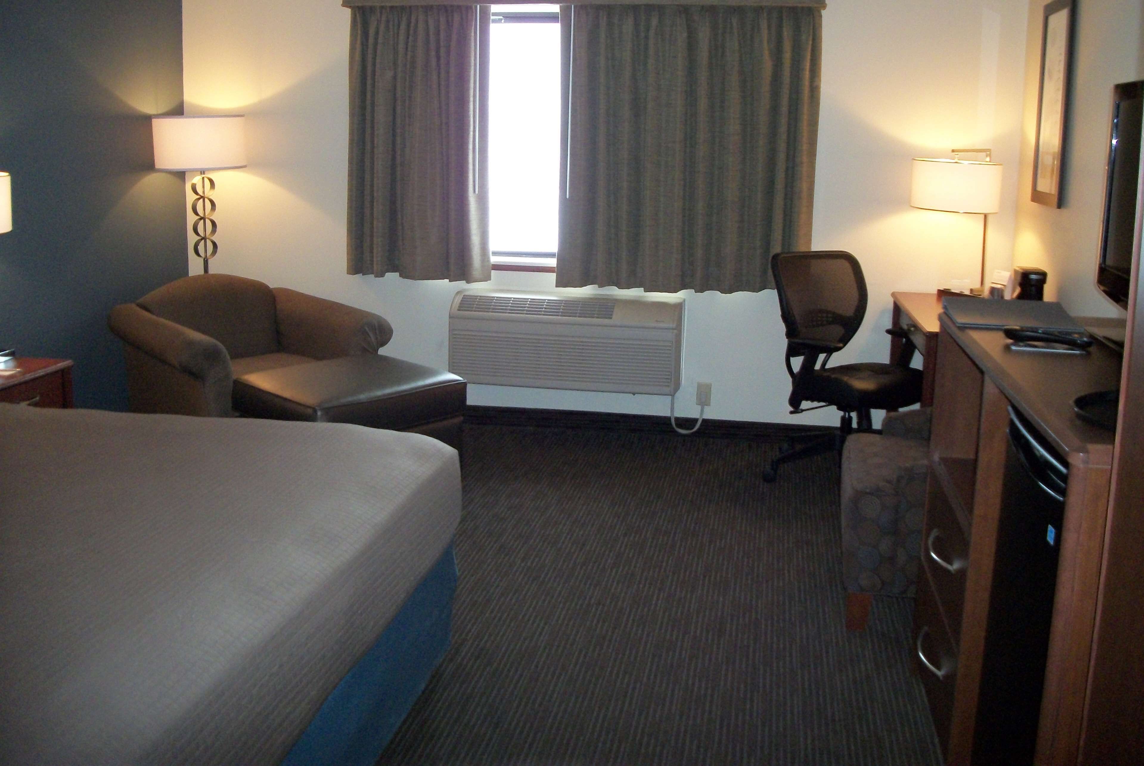 Quarto de hotel com cama, escrivaninha, cadeira, ar condicionado e janela com cortinas.