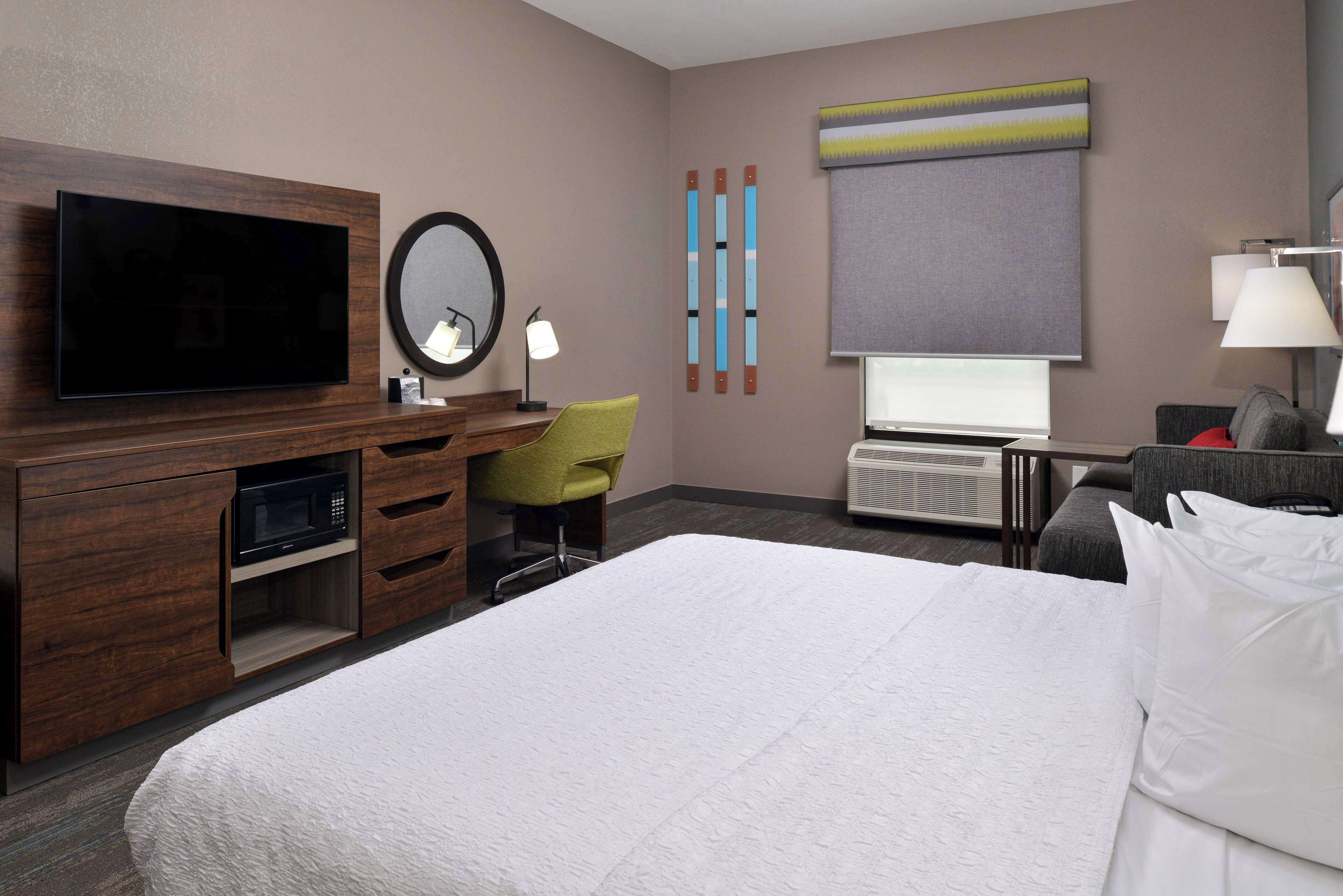 Quarto de hotel com uma cama grande, uma escrivaninha com televisão de tela plana, luminária de mesa, cadeira e janela com persianas.