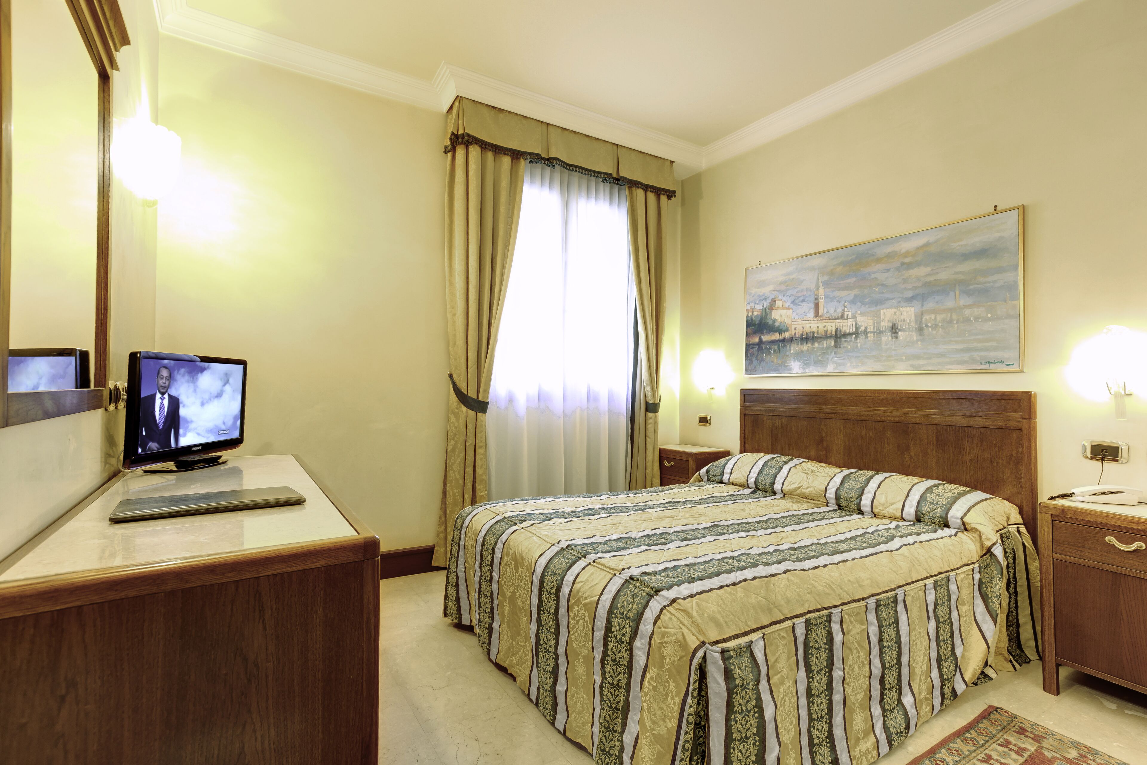 Quarto de hotel com cama, escrivaninha com computador, quadro na parede e janela com cortinas.